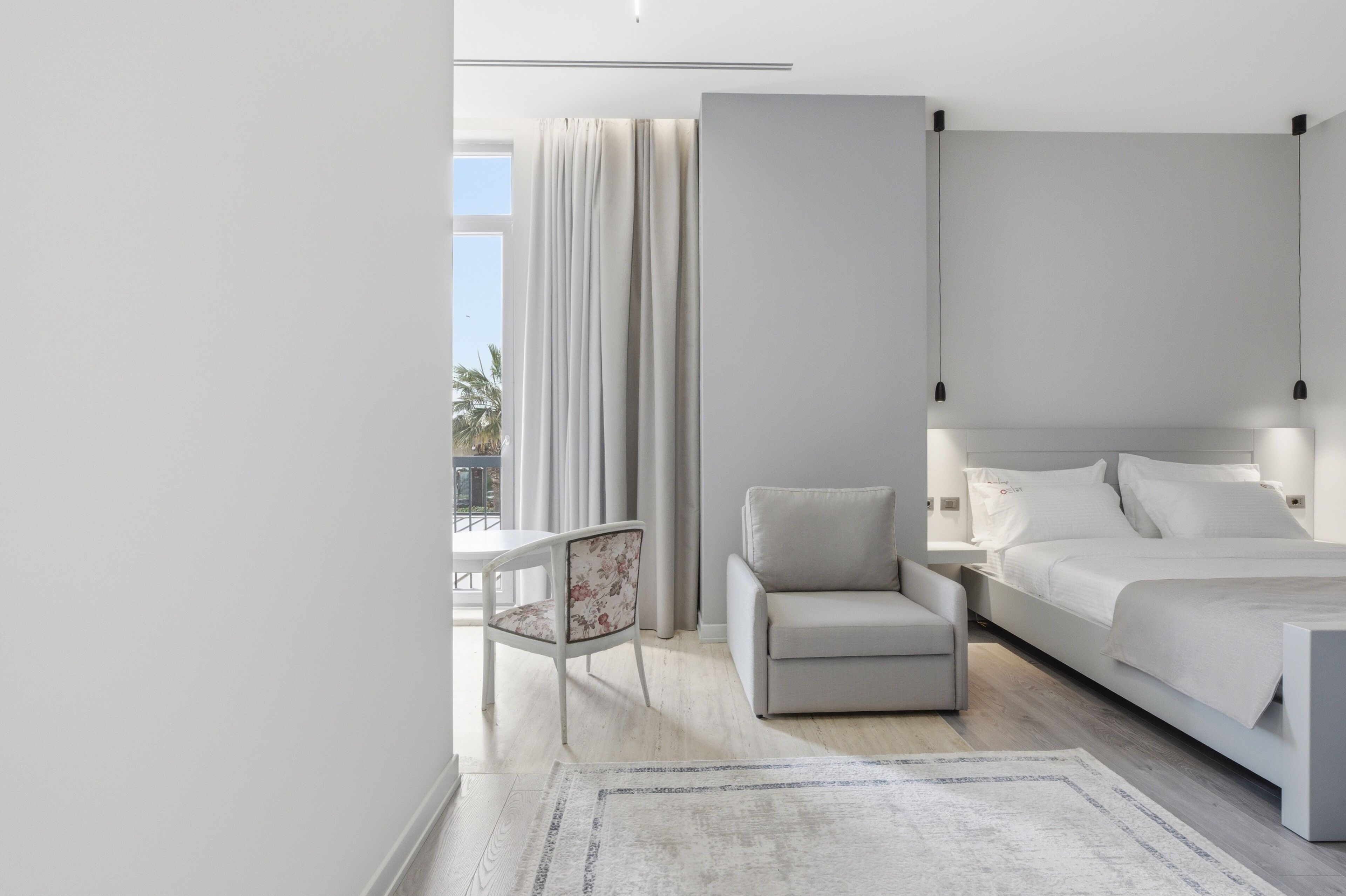 Quarto de hotel moderno com uma cama grande, uma cadeira, uma mesa de cabeceira e vista para o exterior.