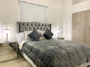 Quarto com cabeceira estofada, cama grande com colcha cinza, duas luminárias de cabeceira e um guarda-roupa.