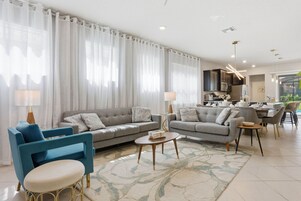 Sala de estar moderna com sofá, cadeiras, mesa de centro e área de cozinha ao fundo.