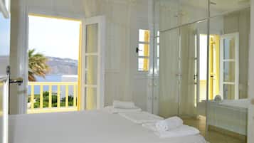 Quarto de hotel com varanda, cama com roupa de cama branca, chuveiro em cabine de vidro e vista para o mar.