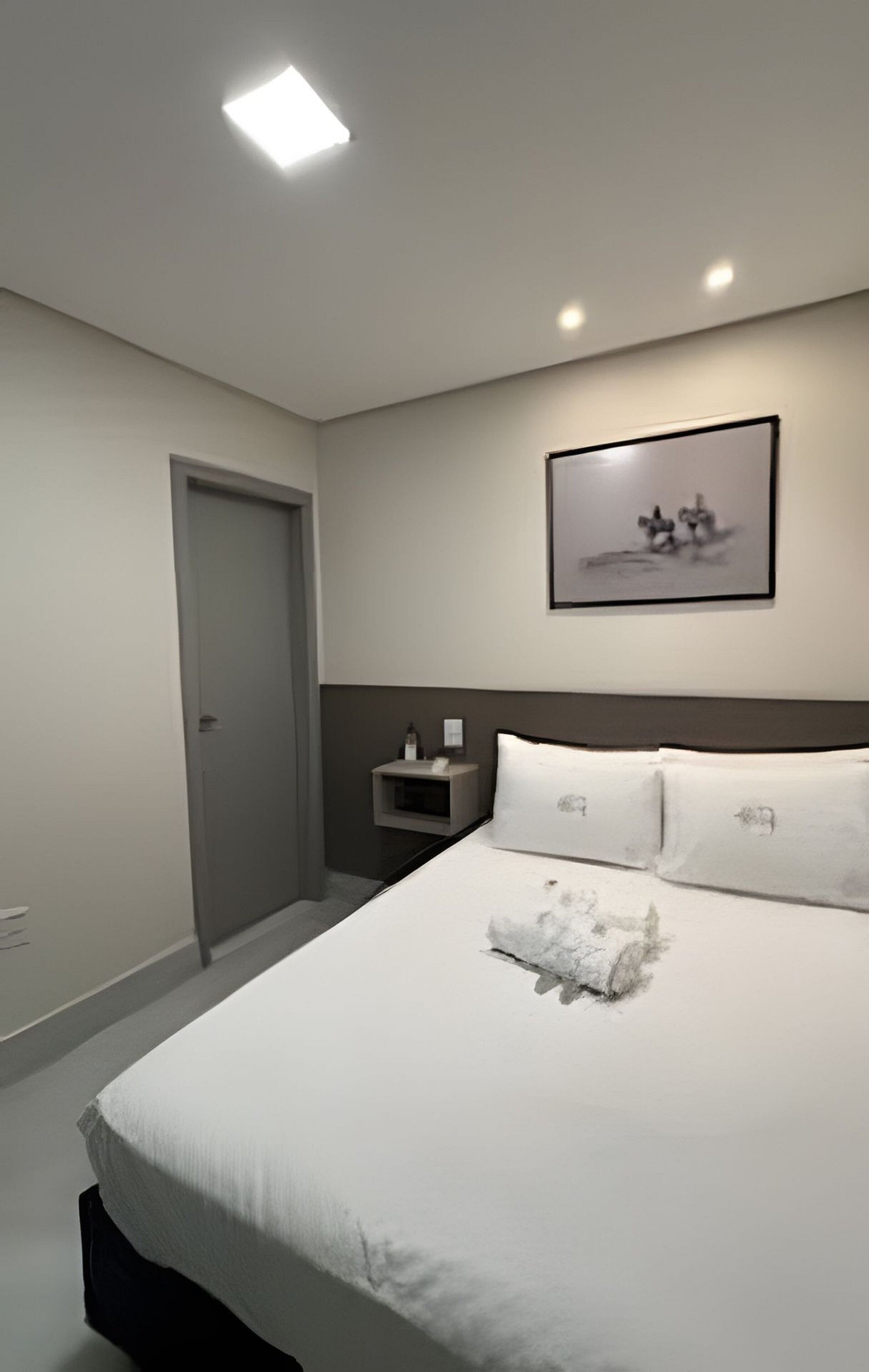Quarto de hotel com uma cama, mesa de cabeceira, quadro na parede e uma porta.