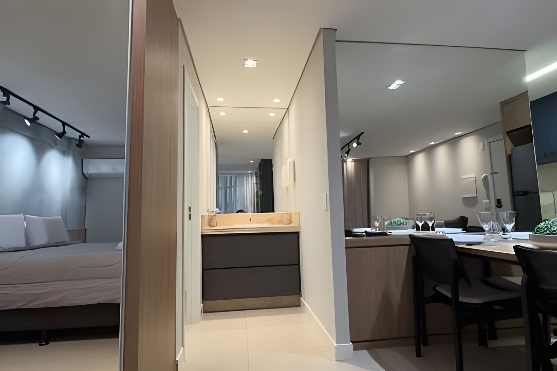 Quarto de hotel moderno com uma cama, uma kitchenette compacta, área de refeições com mesa e cadeiras, e um espelho grande.