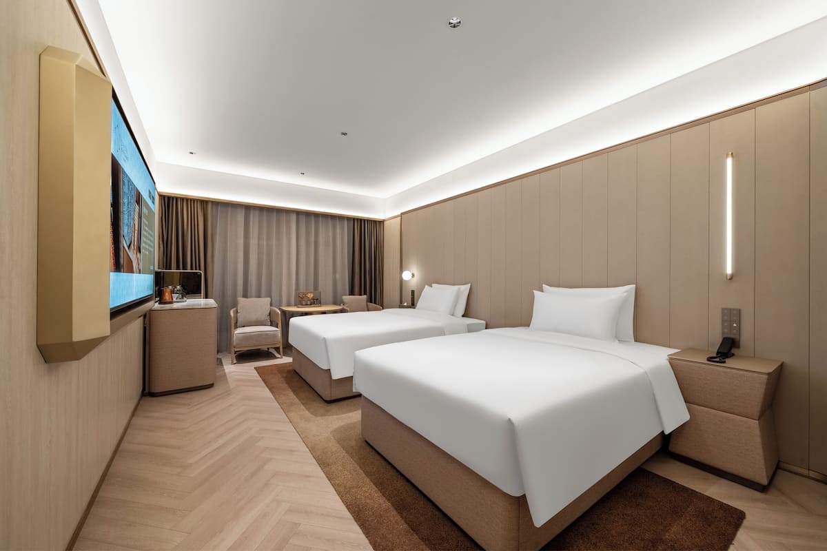 Quarto de hotel moderno com duas camas, uma escrivaninha, uma cadeira, uma televisão e uma janela grande.
