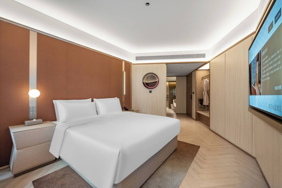 Quarto de hotel moderno com uma cama grande, uma mesa de cabeceira com luminária, uma TV fixada na parede e piso de madeira.