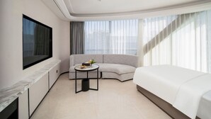 Quarto de hotel moderno com sofá curvo, cama, televisão fixada na parede e uma grande janela com cortinas.