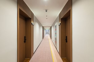 Um corredor longo e estreito com portas de madeira em ambos os lados, um caminho central forrado com carpete e um teto com iluminação embutida.