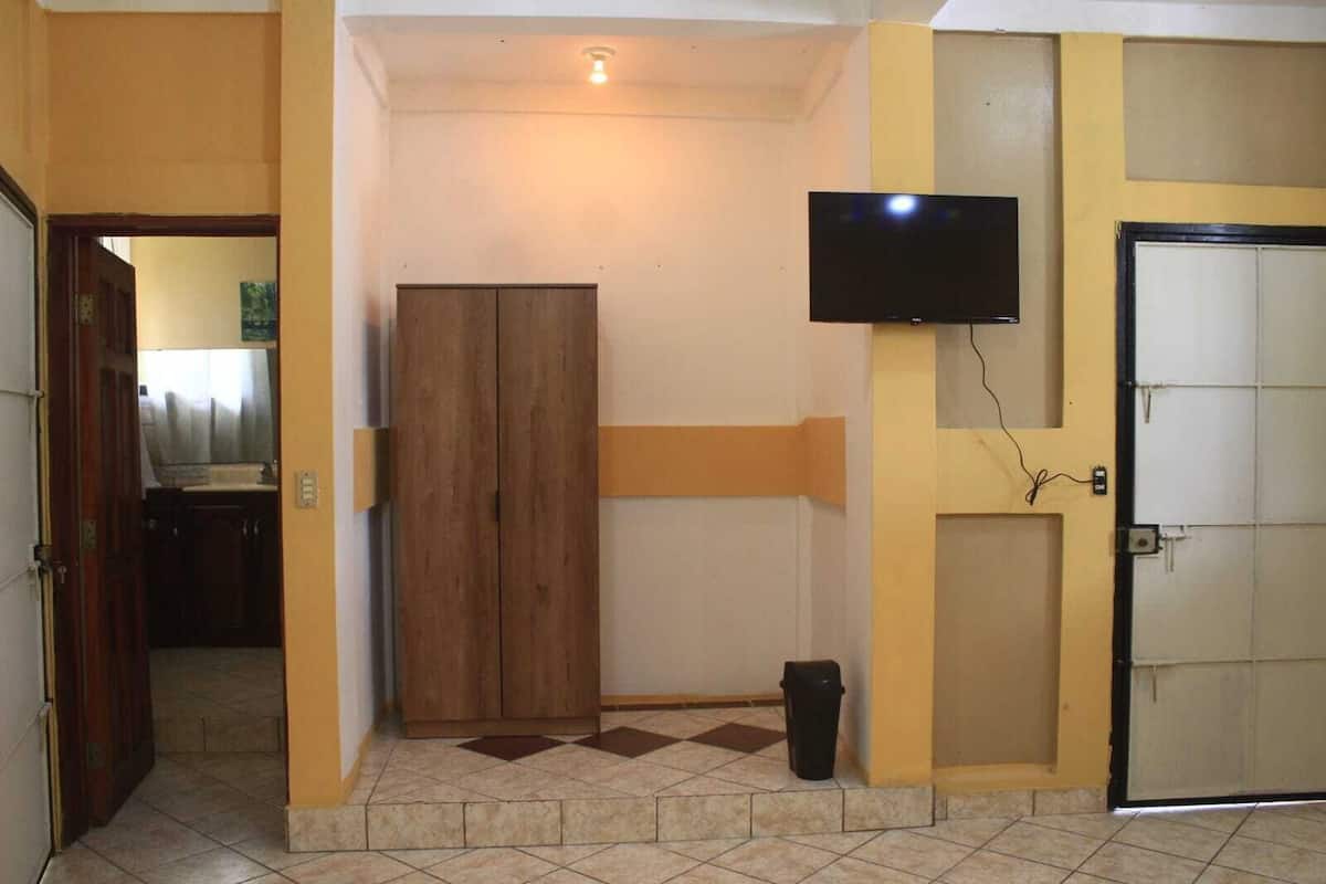 Quarto com porta de madeira, televisão fixada na parede, lixeira e chão revestido de azulejos.