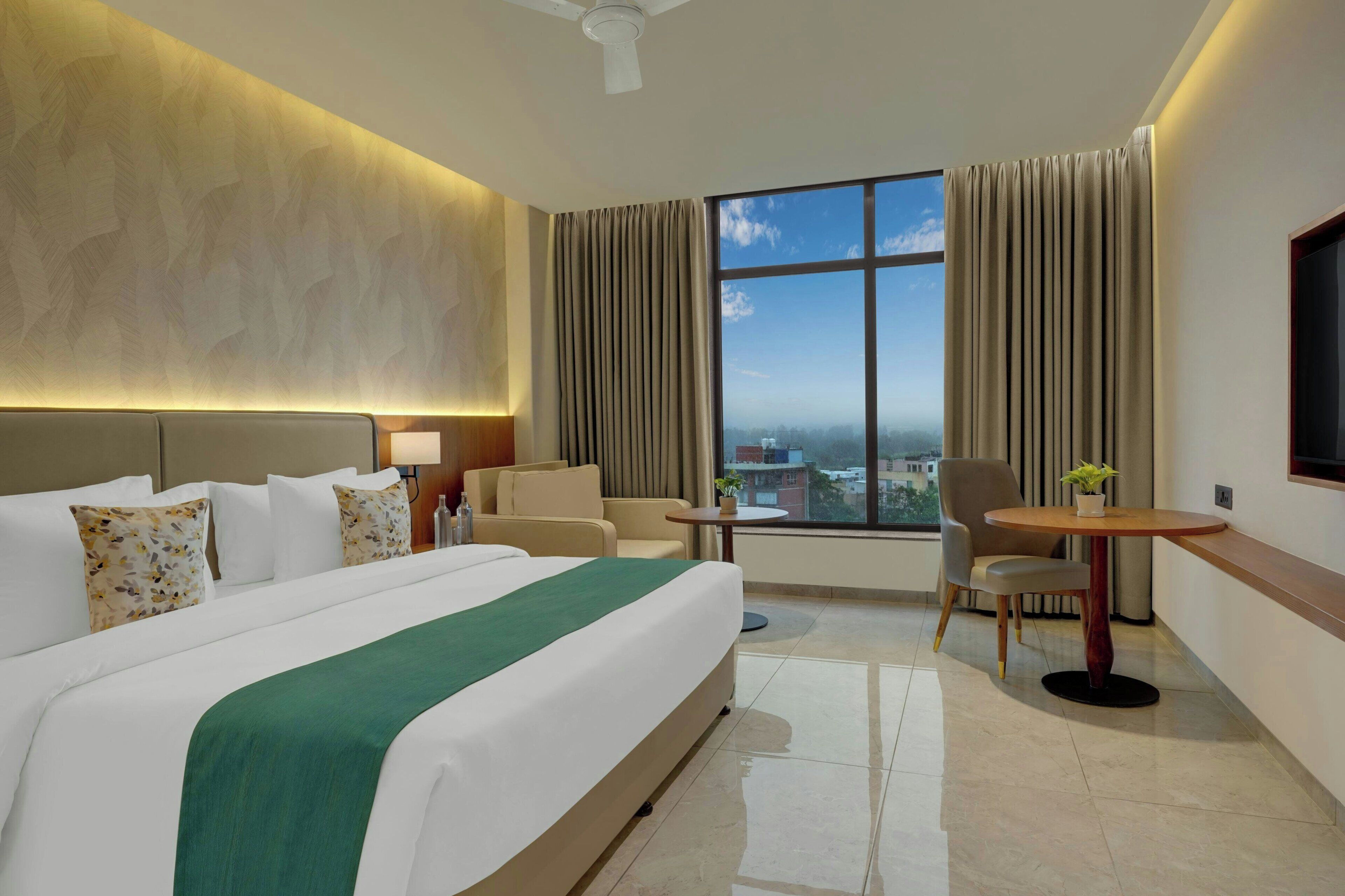 Quarto de hotel com uma cama grande, área de estar com mesa e cadeiras, vista da cidade por uma janela ampla e um ventilador de teto.