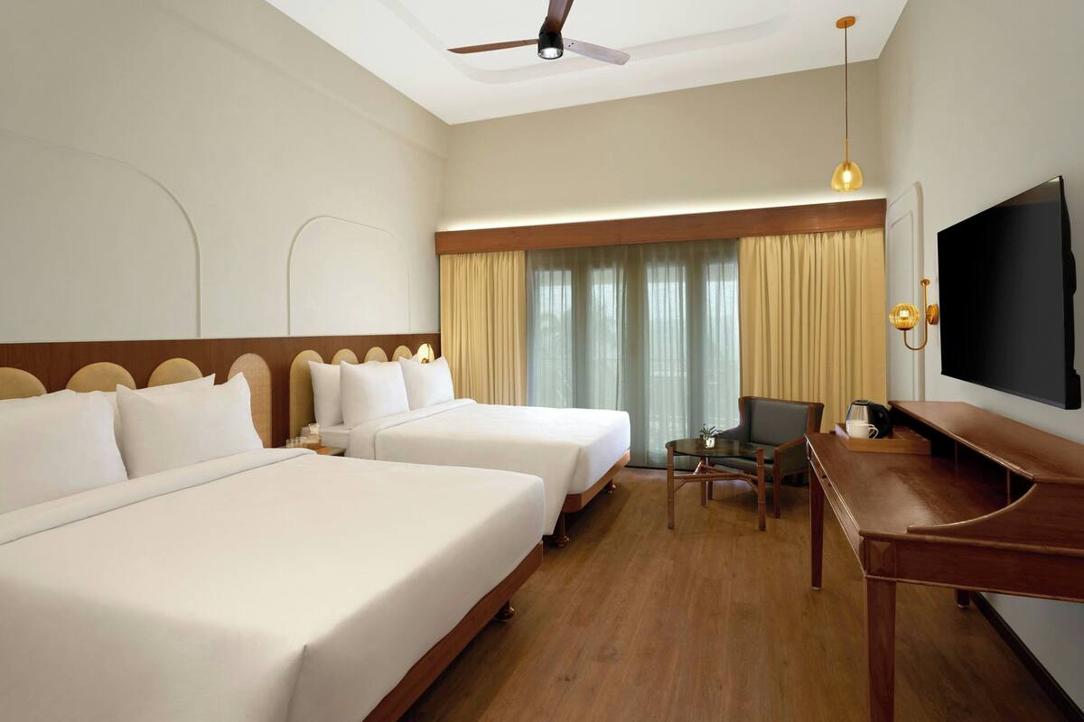 Quarto de hotel com duas camas, uma escrivaninha, uma cadeira, uma televisão e um ventilador de teto.