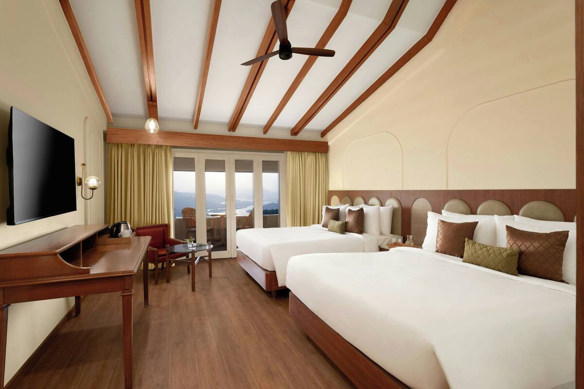 Quarto de hotel com duas camas, televisão, escrivaninha, cadeira e vista para o exterior.