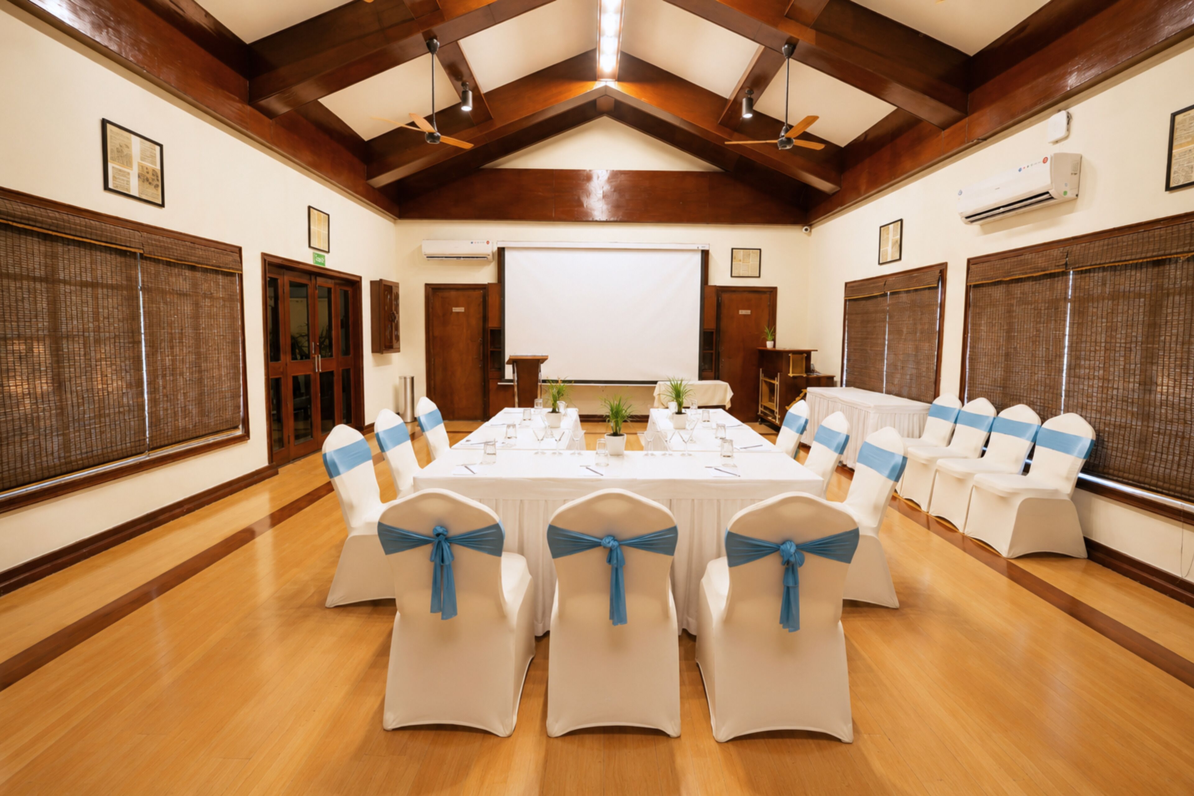 Uma sala de conferências com uma mesa comprida, cadeiras com fitas azuis, piso de madeira e uma tela branca grande.