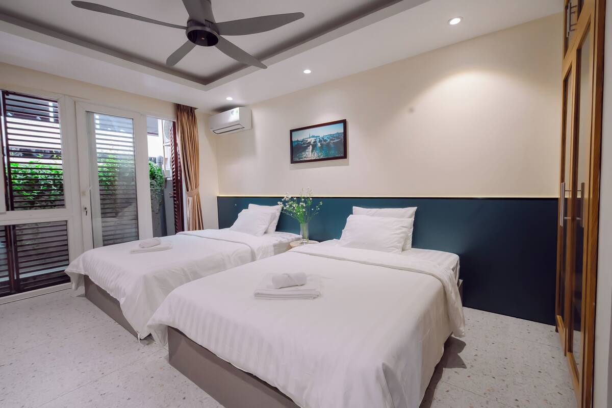 Quarto de hotel com duas camas, ventilador de teto, ar condicionado, um quadro pendurado na parede e uma porta de vidro deslizante.