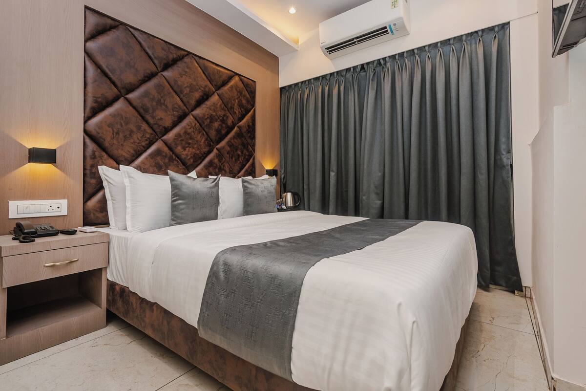Quarto de hotel com uma cama grande, uma mesinha de cabeceira, ar condicionado fixado na parede e uma cortina.