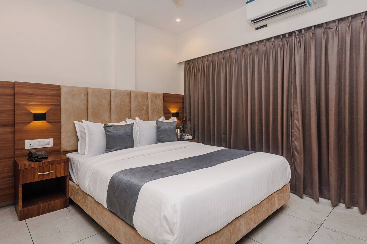 Quarto de hotel moderno com uma cama grande, cabeceira de madeira, abajures e uma janela com cortinas.