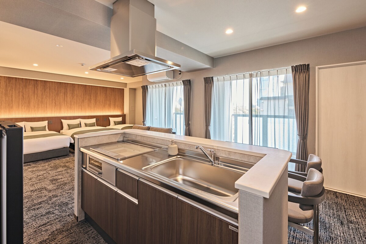 Quarto de hotel moderno com cozinha compacta, uma janela grande com cortinas, cama com almofadas e piso coberto por carpete.