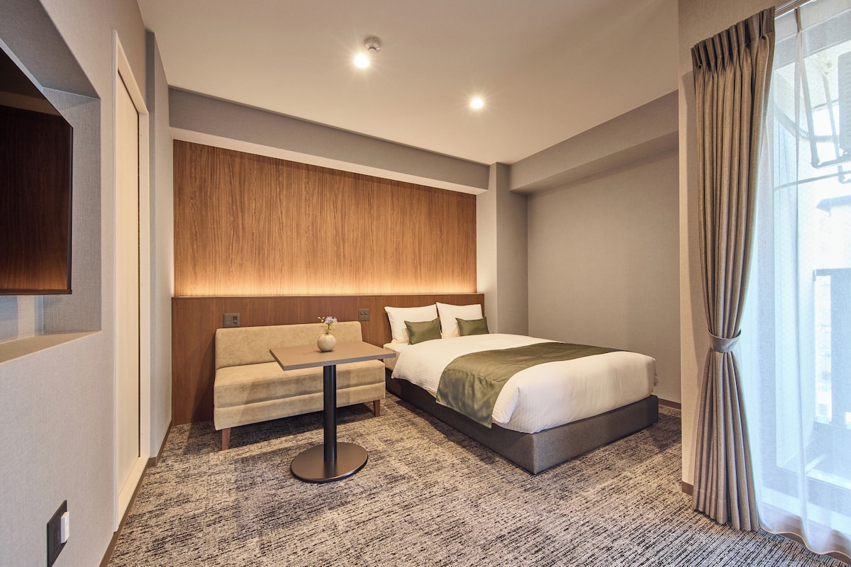 Quarto de hotel com cama, sofá, mesa pequena e televisão presa à parede.
