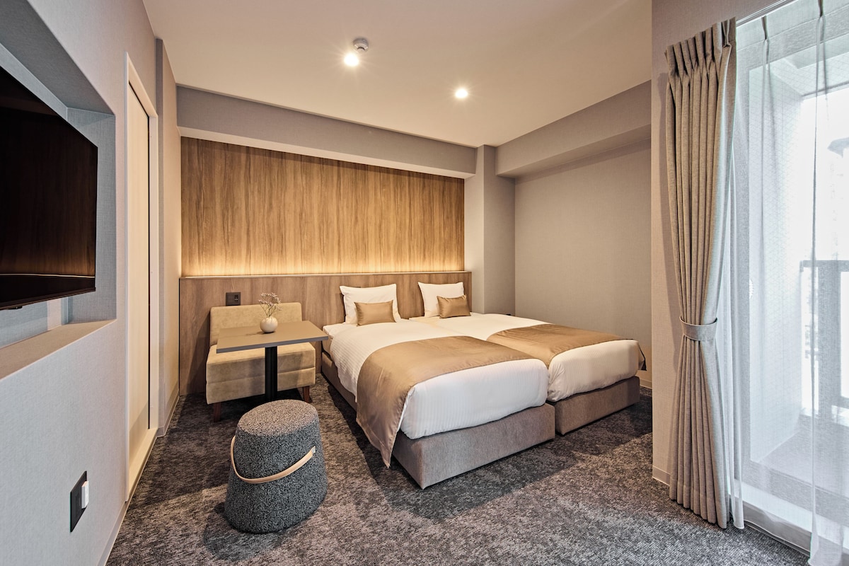 Quarto de hotel moderno com uma cama grande, mesa de cabeceira, televisão e janela com cortinas.