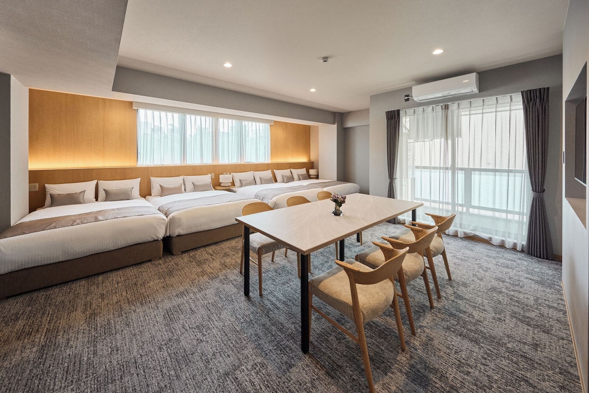 Quarto de hotel com uma cama grande, mesa de jantar com cadeiras, pequena cozinha e uma janela ampla com cortinas.
