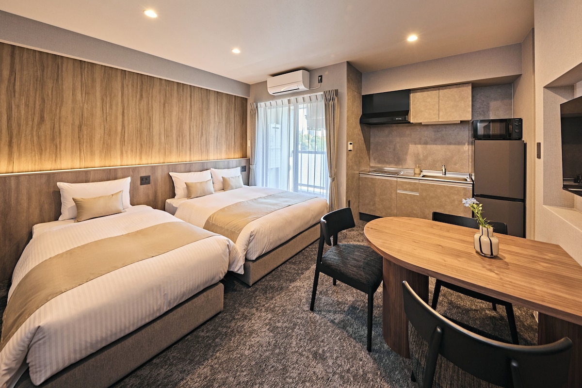 Quarto de hotel com duas camas, uma mesa de jantar, uma área de cozinha e uma janela grande com cortinas.