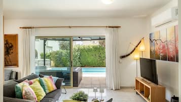 Uma sala de estar com sofá, TV e porta de vidro deslizante que dá para uma piscina.