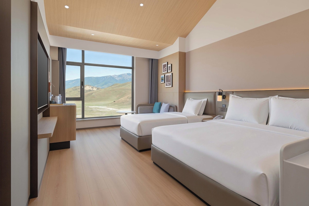 Quarto de hotel com duas camas, uma escrivaninha, televisão e uma grande janela com vista para as montanhas.