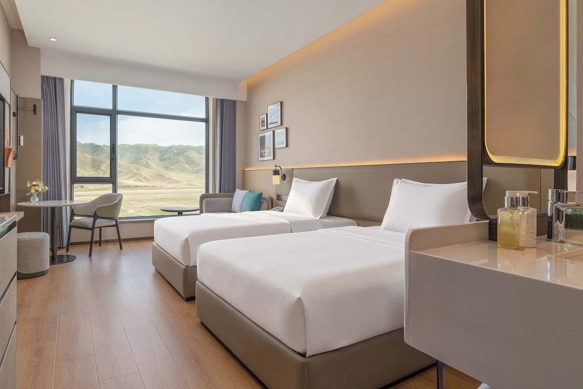 Quarto de hotel com duas camas, uma escrivaninha, uma cadeira, televisão e uma janela grande com vista para as colinas.