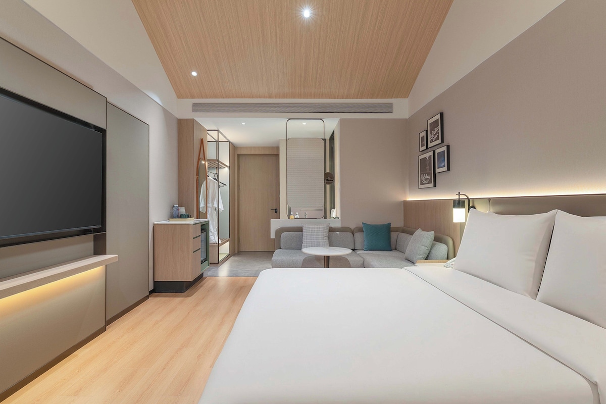 Quarto de hotel moderno com uma cama grande, televisão, área de estar com mesa e um guarda-roupa.
