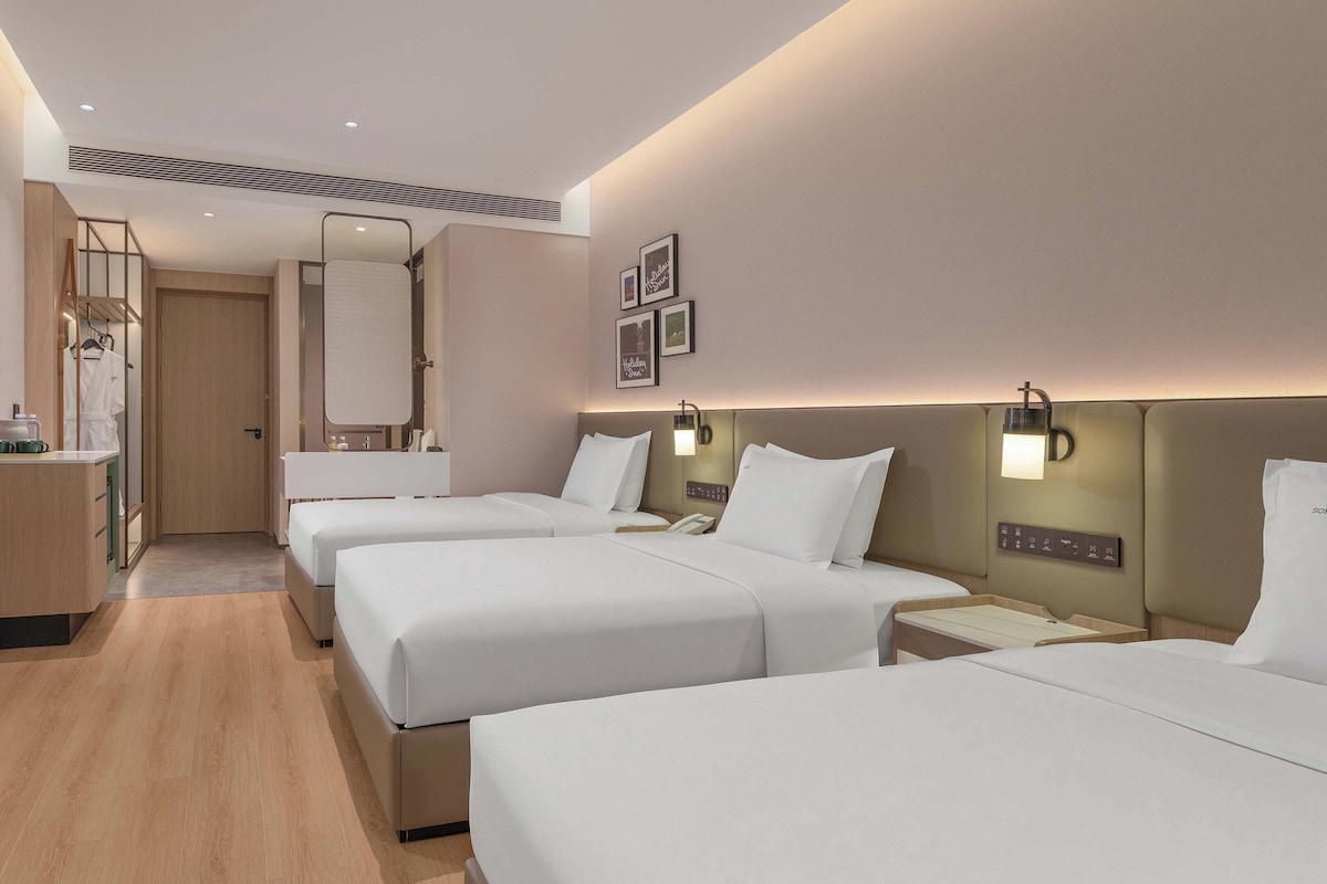 Quarto de hotel com três camas, televisão, banheiro e corredor.