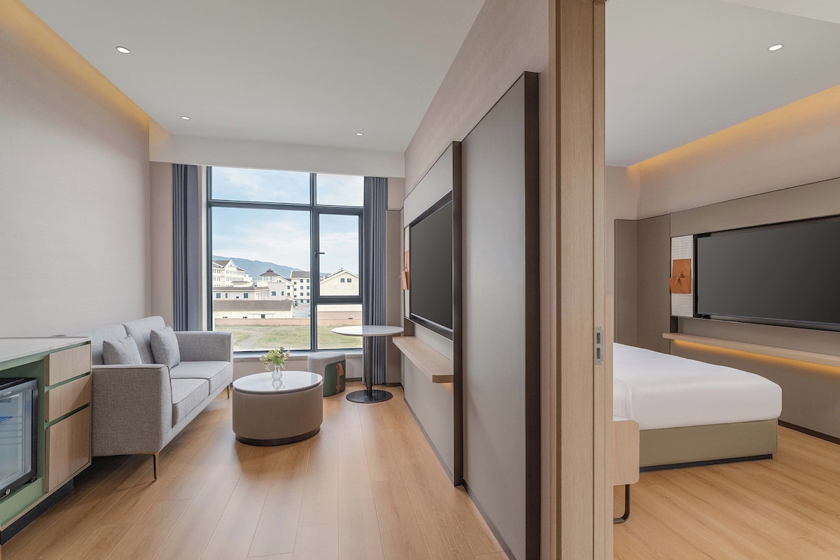 Quarto de hotel moderno com sofá, mesa redonda, cama, televisão e vista para edifícios.