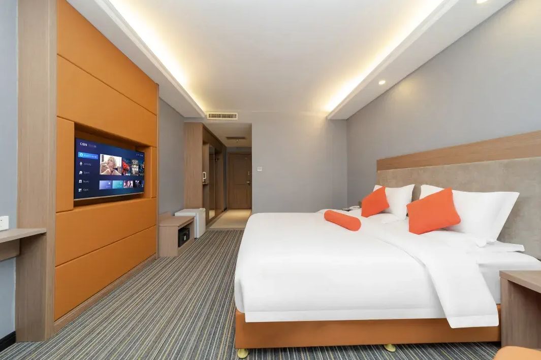 Quarto de hotel com cama, televisão fixada na parede e piso coberto por carpete.