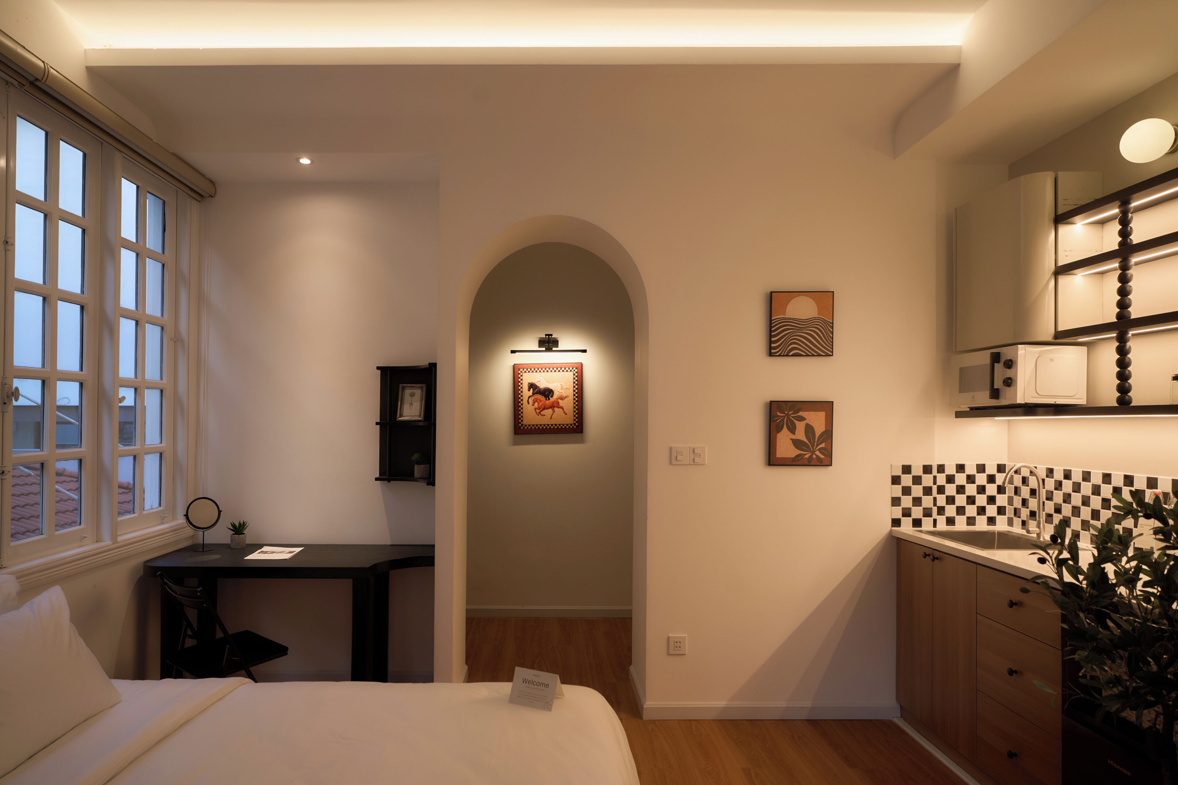 Quarto de hotel com uma cama, uma escrivaninha, uma mesinha, um espelho, um vaso de plantas e vista para o exterior pela janela.