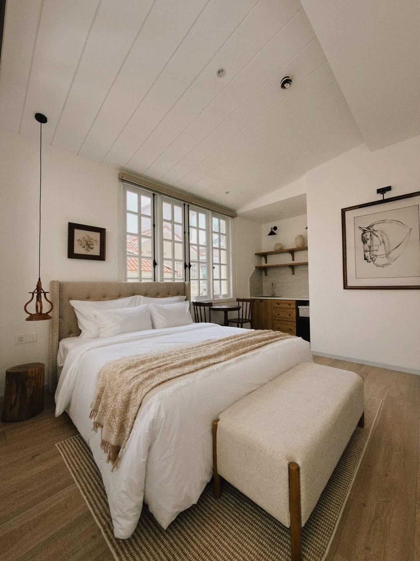 Quarto com uma cama grande, um banco, uma escrivaninha e uma janela.