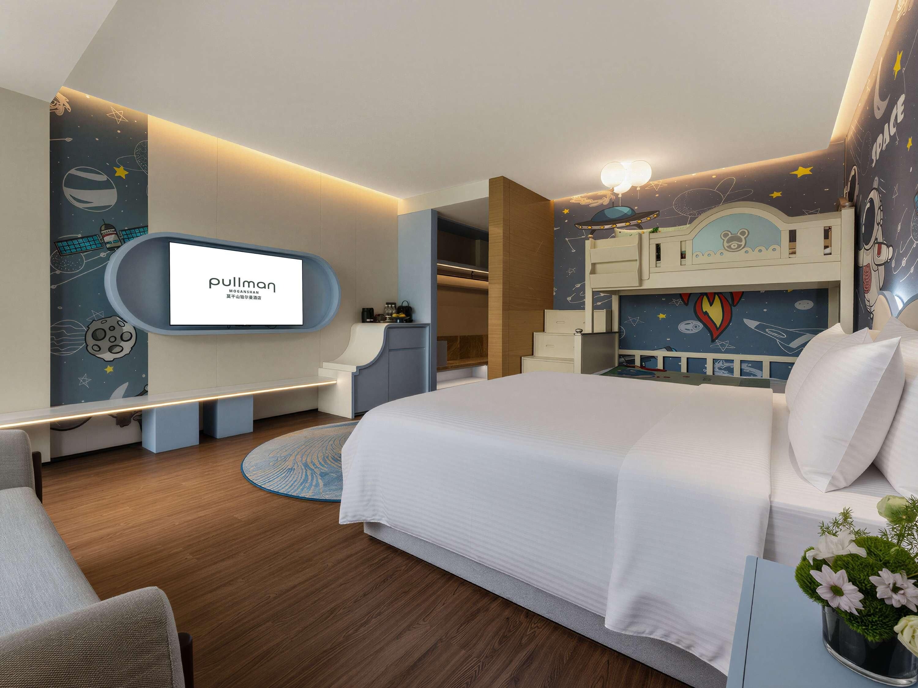 Quarto de hotel moderno com uma cama grande, TV fixada na parede e beliche com decoração temática espacial.