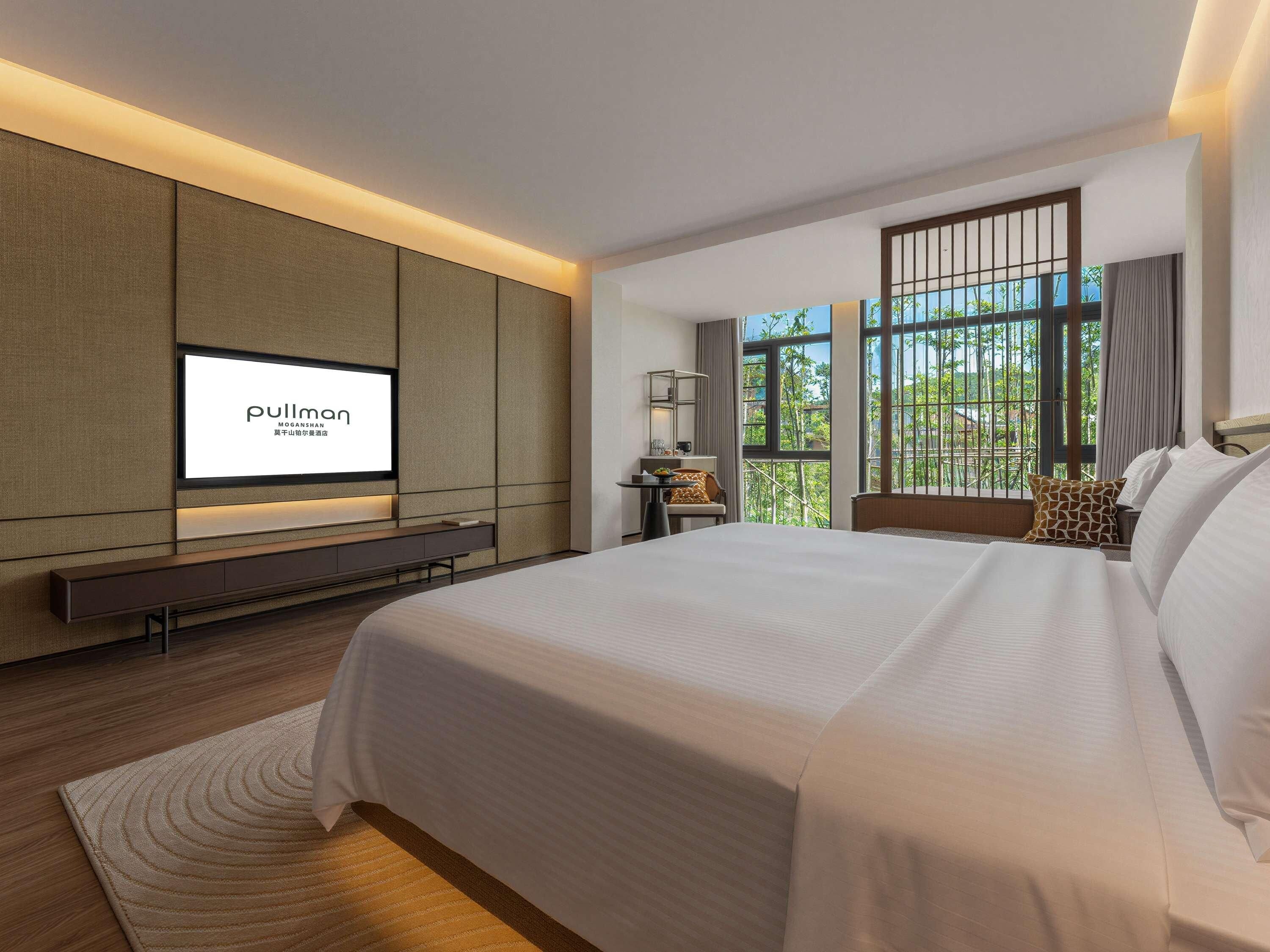 Quarto de hotel moderno com uma cama grande, televisão de tela plana na parede, uma mesa pequena com uma cadeira e uma janela com vista para vegetação.