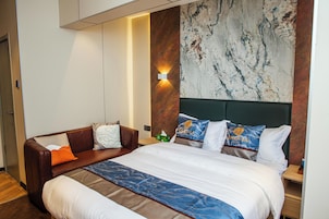 Quarto de hotel com uma cama grande, um sofá de couro marrom, uma parede de mármore e uma mesa de cabeceira com abajur.