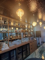 Um bar com prateleiras cheias de garrafas e copos, bancada de mármore e luminárias pendentes.