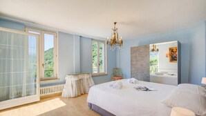 Quarto com cama grande, lustre, janela com vista, radiador e uma mesinha.