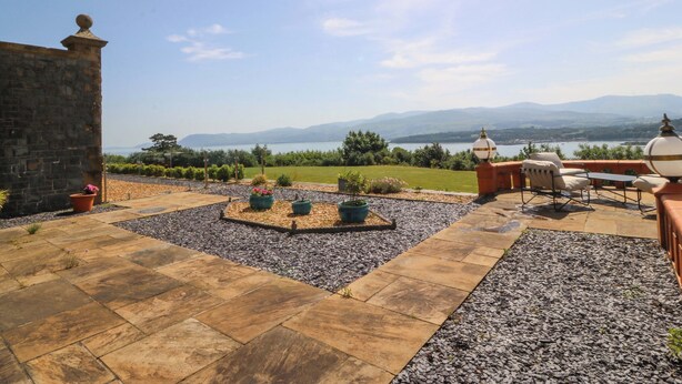 Um pátio com piso de pedra, plantas em vasos, móveis de exterior e vista para um lago e montanhas.