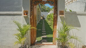 Um caminho estreito que leva a uma porta com trabalhos em madeira detalhados, ladeado por vasos de plantas, e uma visão clara de uma escadaria e vegetação ao fundo.