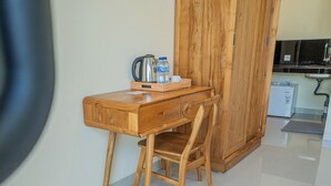 Uma secretária de madeira com uma cadeira, uma chaleira e garrafas de água num quarto com soalho em azulejos e um guarda-roupa de madeira.