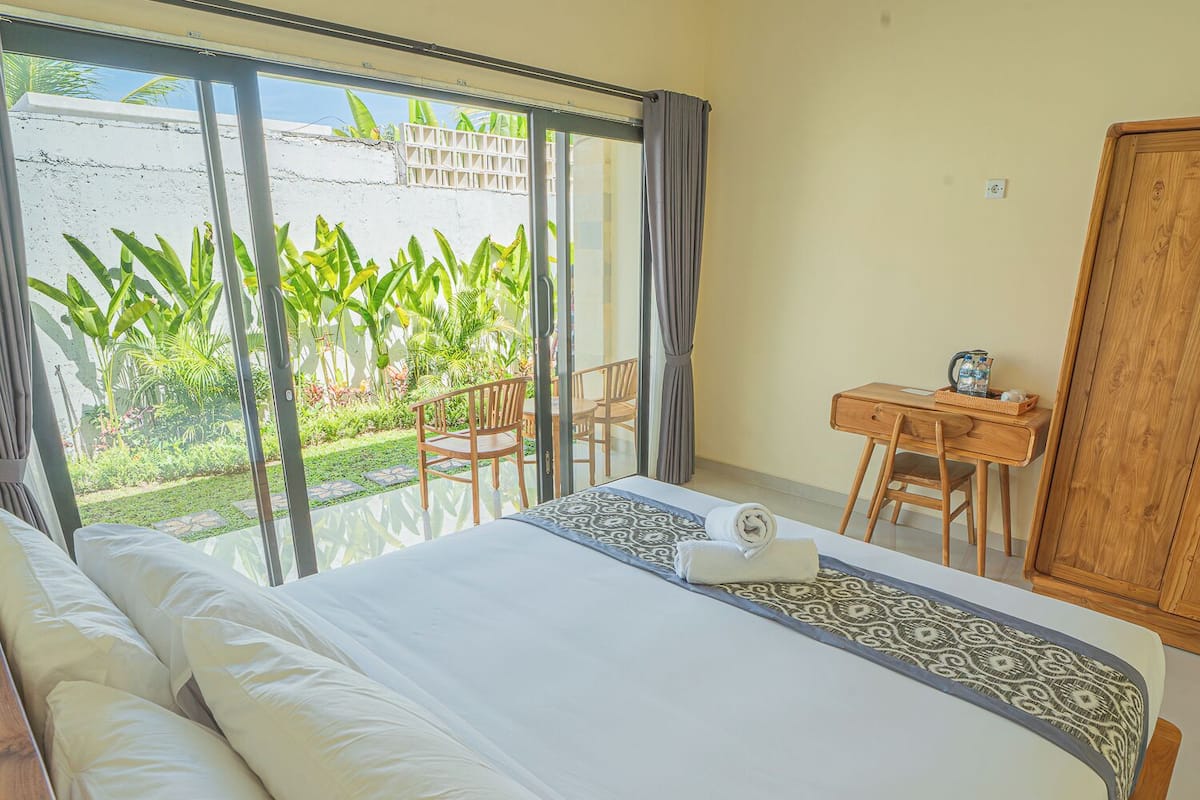 Quarto de hotel com uma cama grande, uma escrivaninha de madeira, uma cadeira e vista para um jardim através de uma porta de correr.