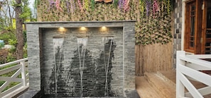 Uma fonte de pedra com três bicas, cerca de madeira e um muro com plantas pendentes.