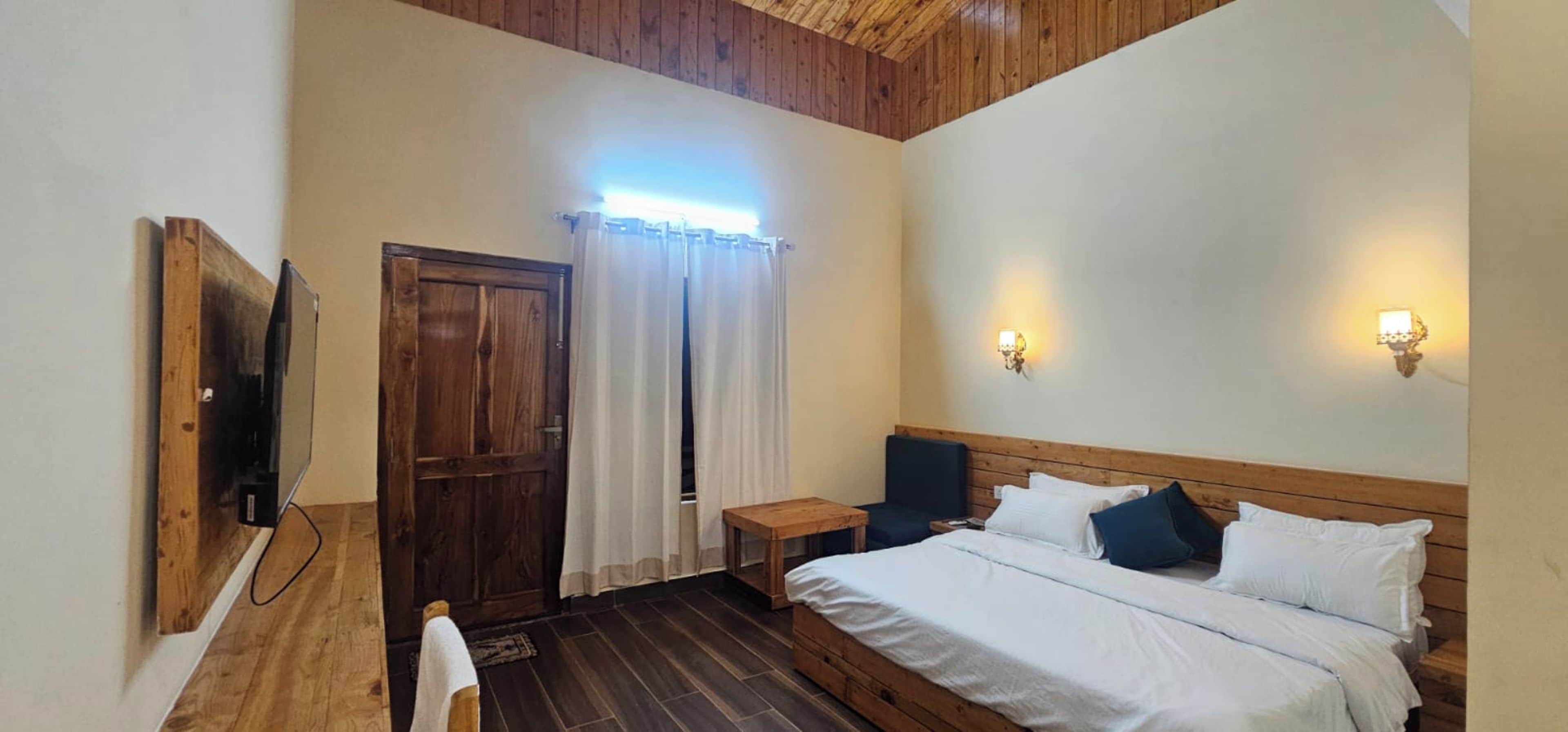 Quarto com teto de madeira, cama com roupa de cama branca, mesa de cabeceira, cadeira e televisão.