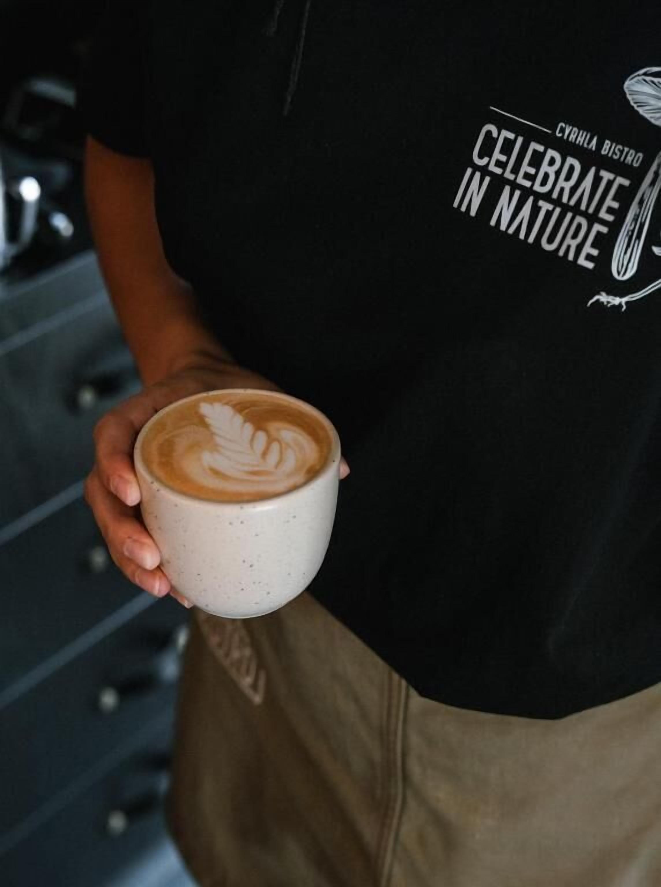 Uma pessoa segurando uma xícara de café com arte de latte, vestindo uma camisa preta com um logotipo e texto.