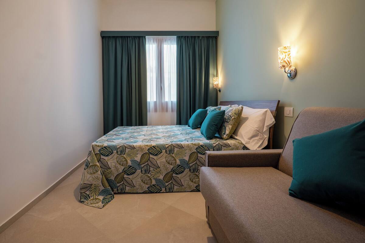 Um quarto com cama, sofá, janela com cortinas e luminárias fixadas na parede.