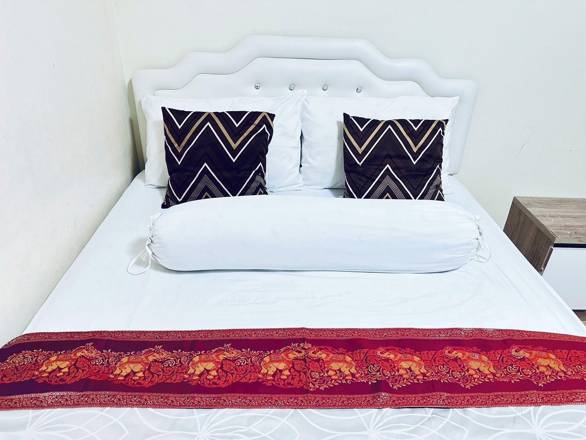Uma cama com cabeceira branca, duas almofadas estampadas, uma almofada branca e uma borda decorativa vermelha.