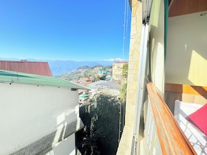 Uma varanda estreita com vista para uma paisagem montanhosa, uma parede de pedra e um edifício vermelho ao longe.