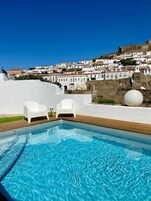 Piscina com duas cadeiras brancas, uma esfera branca grande e vista para uma encosta com edifícios.