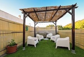 Uma área de estar coberta ao ar livre com grama sintética, cadeiras brancas e uma estrutura de pérgola.