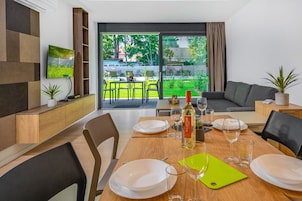 Uma área de refeições moderna com uma mesa de madeira posta para dois, um sofá e vista para um jardim através de uma porta de vidro.